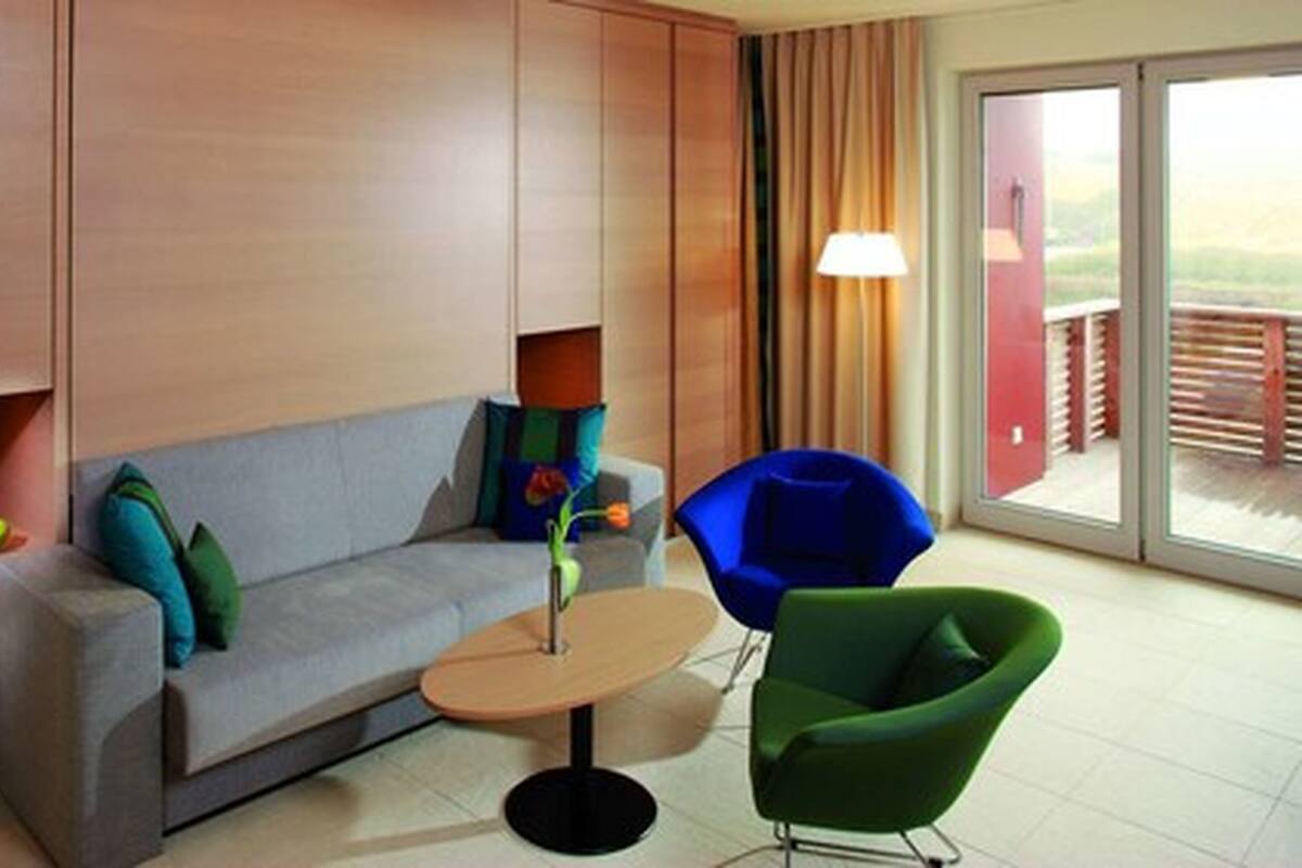 Quarto de hotel moderno com sofá cinzento, mesa redonda, poltrona verde e uma grande janela com vista.