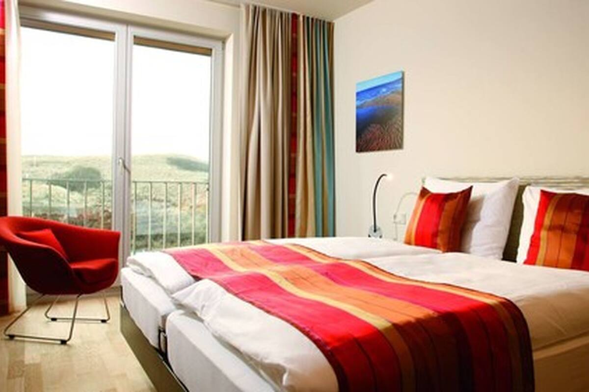 Quarto de hotel com uma cama grande, uma cadeira vermelha, uma varanda com vista e um quadro na parede.