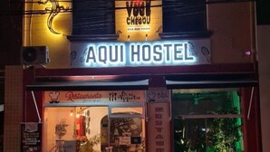 Um hostel com uma placa bem iluminada com a inscrição 'AQUI HOSTEL' e uma placa de restaurante abaixo.