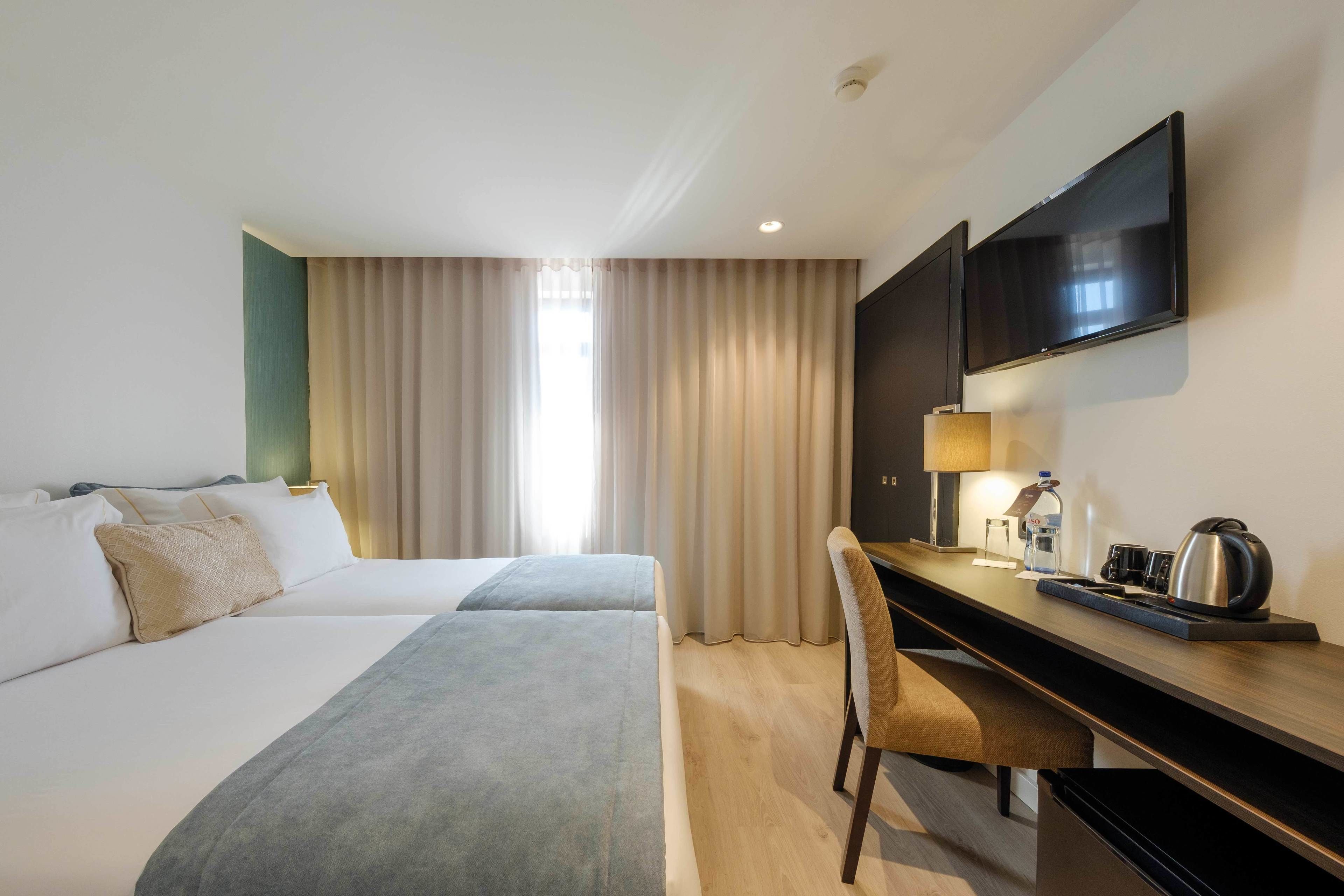 Quarto de hotel com duas camas, uma mesa, uma cadeira, uma televisão e uma janela com cortinas.