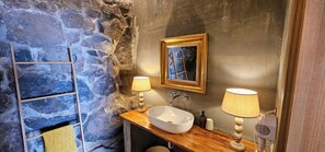 Um banheiro com parede de pedra, toucador de madeira, pia branca, duas luminárias fixadas na parede e espelho emoldurado.