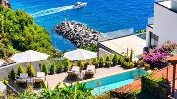Piscina no terraço com espreguiçadeiras e guarda-sóis, com vista para um litoral rochoso e o mar.