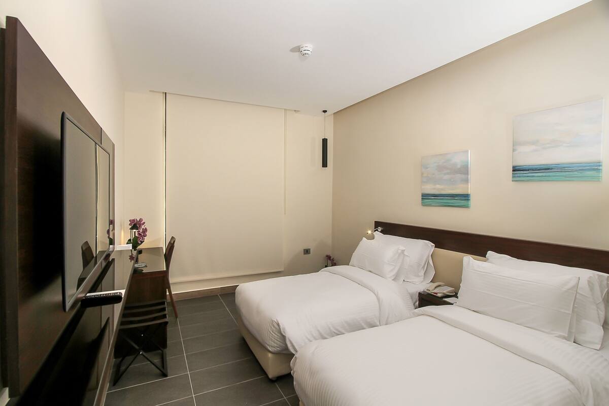 Quarto de hotel com duas camas, uma escrivaninha com cadeira, televisão e obra de arte na parede.