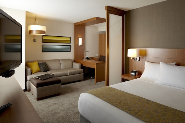 Quarto de hotel com cama, escrivaninha, sofá, televisão e espelho.