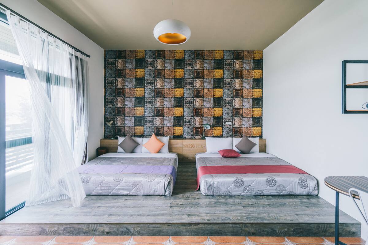 Quarto com duas camas, parede estampada, luminária no teto e janela com cortinas.