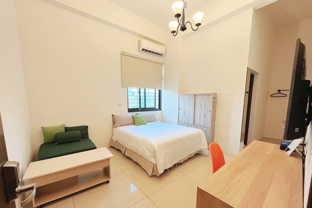 Quarto compacto com cama, escrivaninha, cadeira e um pequeno espaço para sentar.