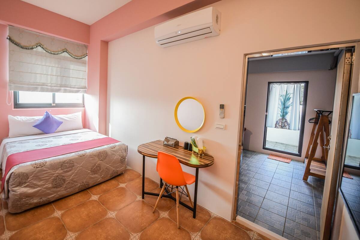 Quarto com cama, uma mesinha com cadeira, ar condicionado e janela com cortina.
