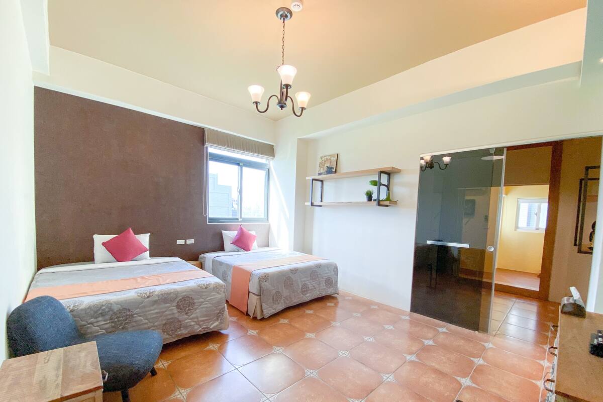 Quarto de hotel com duas camas, um candelabro, janela com cortinas, um pequeno aparador com uma planta e uma porta de vidro deslizante que dá para um corredor.