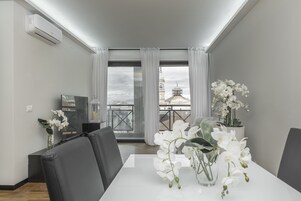 Uma área de jantar moderna com uma mesa branca, cadeiras pretas e orquídeas brancas. Uma grande janela oferece vista para um edifício com uma cúpula e uma varanda.