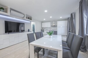 Uma sala de jantar moderna com uma mesa branca e cadeiras pretas, uma televisão de tela plana na parede e um vaso de planta na mesa.