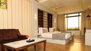 Quarto de hotel com sofá, duas camas, mesa de centro de madeira e uma janela grande com cortinas.