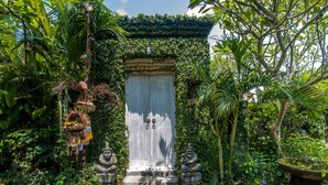 Entrada de um jardim com uma porta branca, duas estátuas de Buda em pedra e vegetação exuberante.