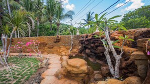 Um jardim ajardinado com muro de pedra, um pequeno lago e diversas plantas, incluindo flores cor-de-rosa.