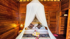 Quarto de madeira com uma cama sob um dossel, almofadas com padrões geométricos e uma mesa de cabeceira com luminária.