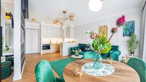 Uma sala de estar moderna com uma mesa de jantar redonda de madeira, sofás de veludo verde e uma cozinha ao fundo.