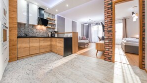 Cozinha moderna com armários de madeira, bancada preta e painel decorativo estampado. Há uma área de refeições com sofá cinza e mesa de centro. O espaço possui parede com detalhes em tijolo e um espelho grande.