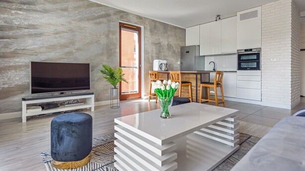 Uma sala de estar moderna com uma TV de tela plana, uma mesa de centro e uma área de jantar com cadeiras, e uma cozinha ao fundo.
