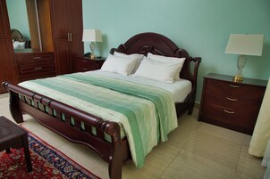 Uma cama bem arrumada com cabeceira de madeira, mesa de cabeceira com abajur e criado-mudo com luminária.