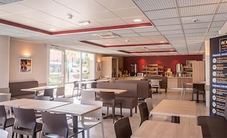 Uma ampla área de refeições interna com várias mesas e cadeiras, um balcão com menu e vista para o exterior por grandes janelas.