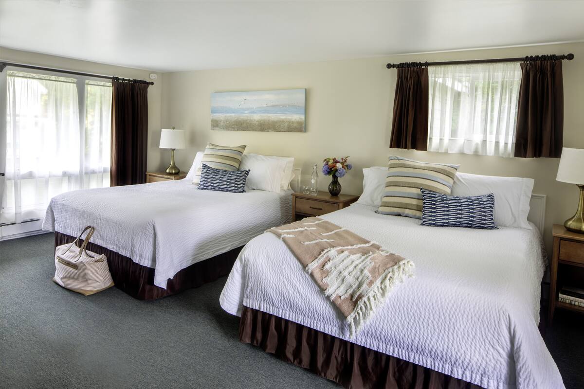 Quarto de hotel com duas camas, uma janela grande com cortinas, um quadro na parede e uma mesa de cabeceira com abajur.