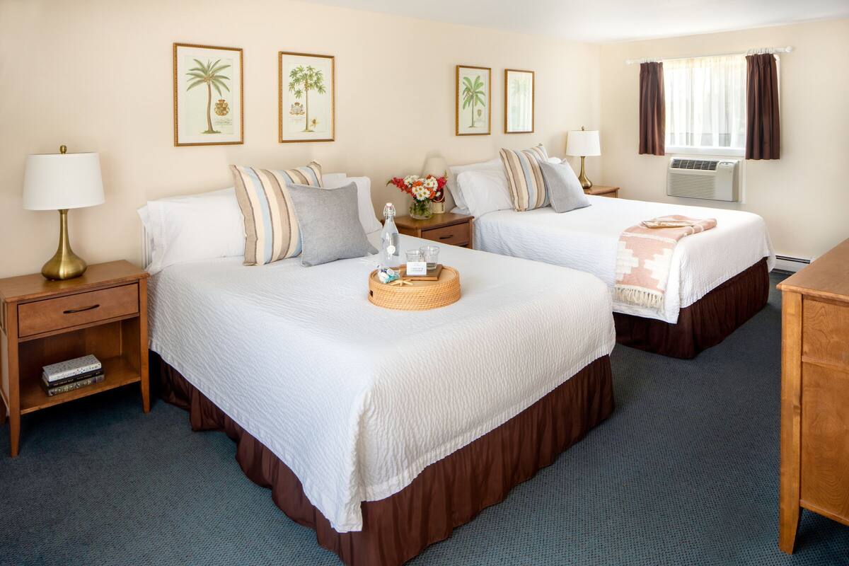 Quarto de hotel com duas camas, uma mesa de cabeceira com abajur, uma pequena estante com livros e quadros nas paredes.