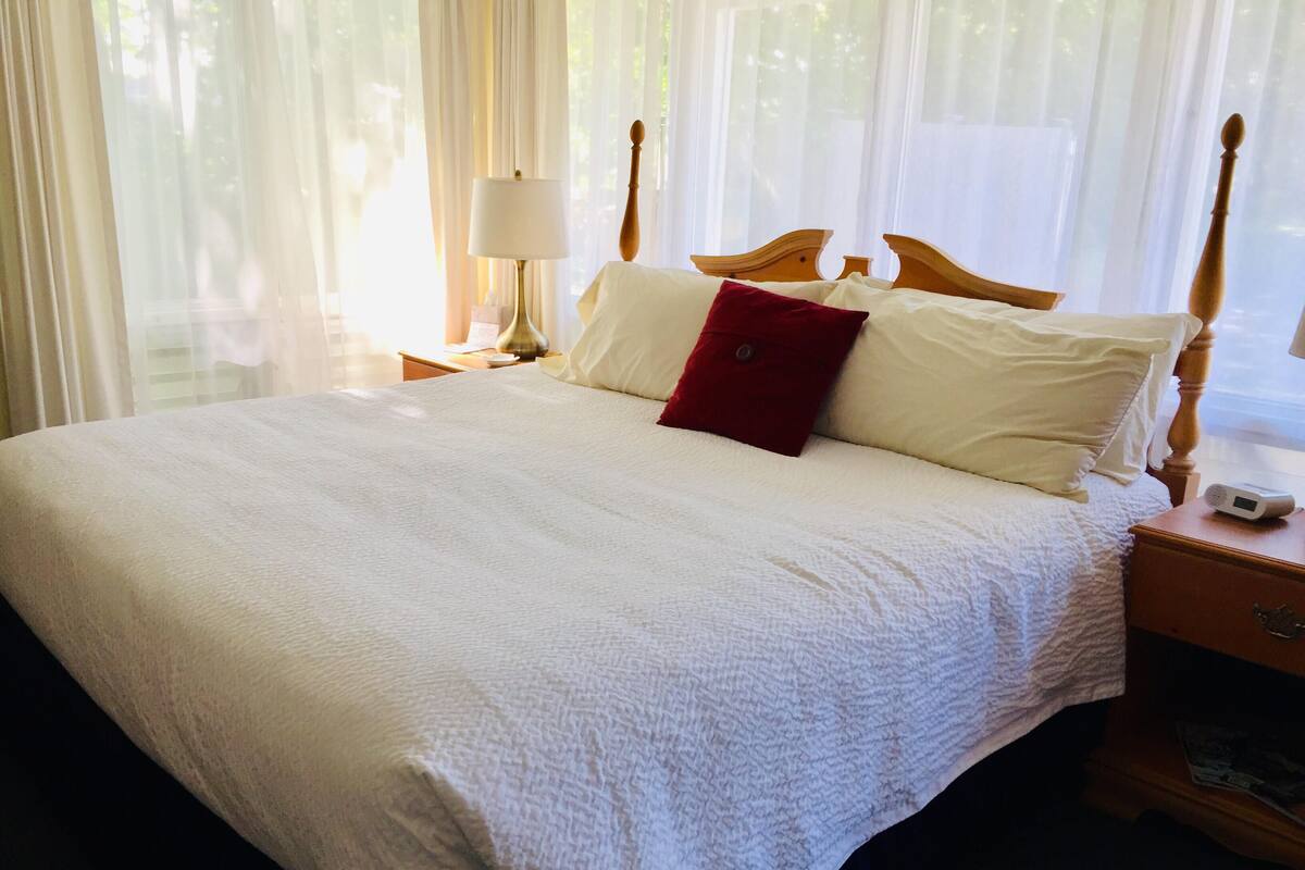 Cama bem arrumada com edredom branco, travesseiro vermelho e cabeceira de madeira. Um criado-mudo com luminária e um pequeno aparelho eletrônico.