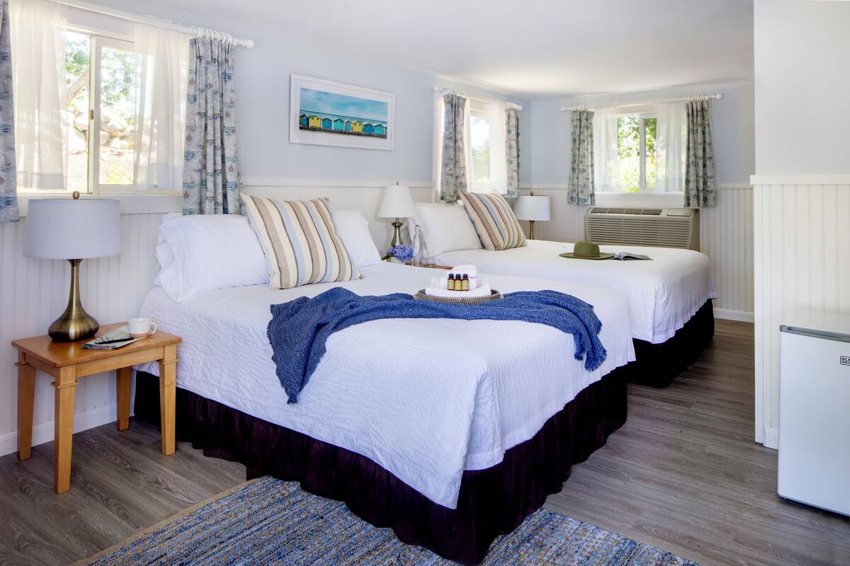 Quarto duplo com duas camas, uma mesinha com abajur, frigorífico e janela com cortinas.