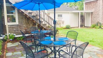 Um pátio com mesa e cadeiras, um guarda-sol azul e uma escadaria que leva a uma casa.