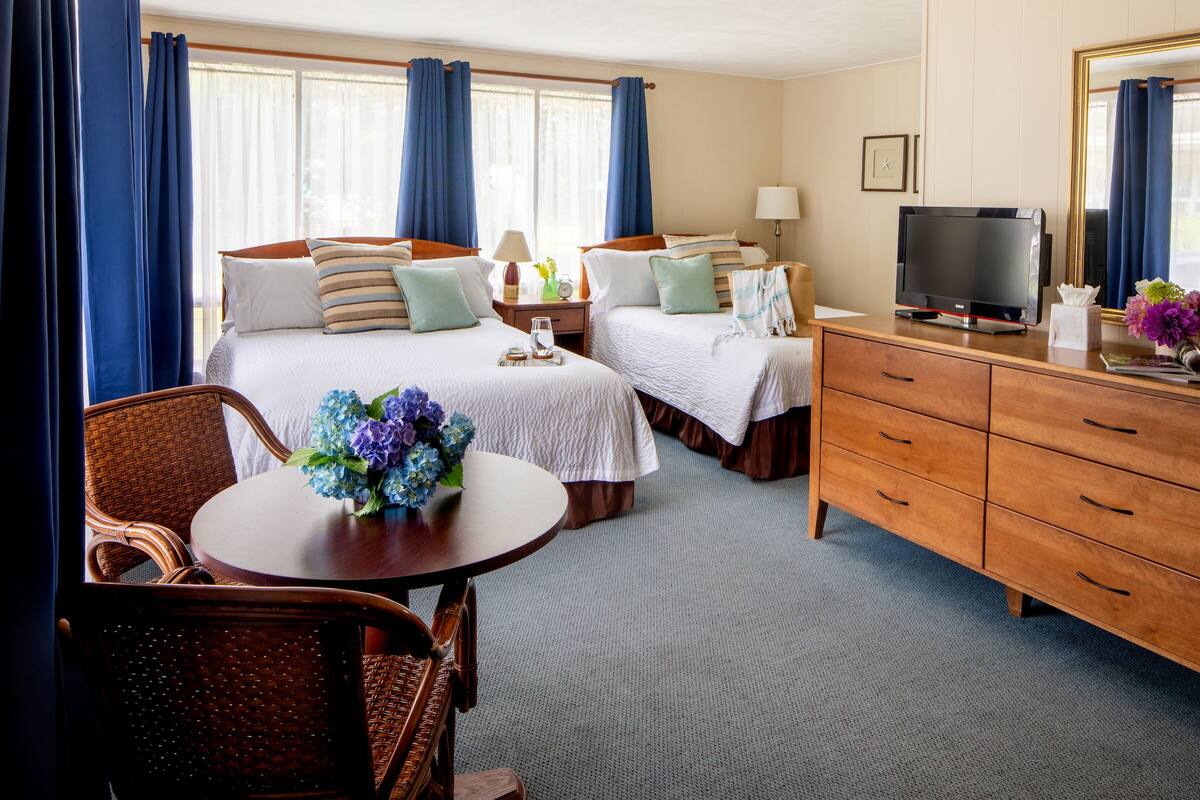 Quarto de hotel com duas camas, televisão, penteadeira, mesa com um vaso de flores e uma cadeira.