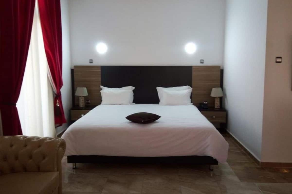 Quarto de hotel com uma cama grande, duas mesas de cabeceira com abajures, uma cortina vermelha e uma poltrona bege.