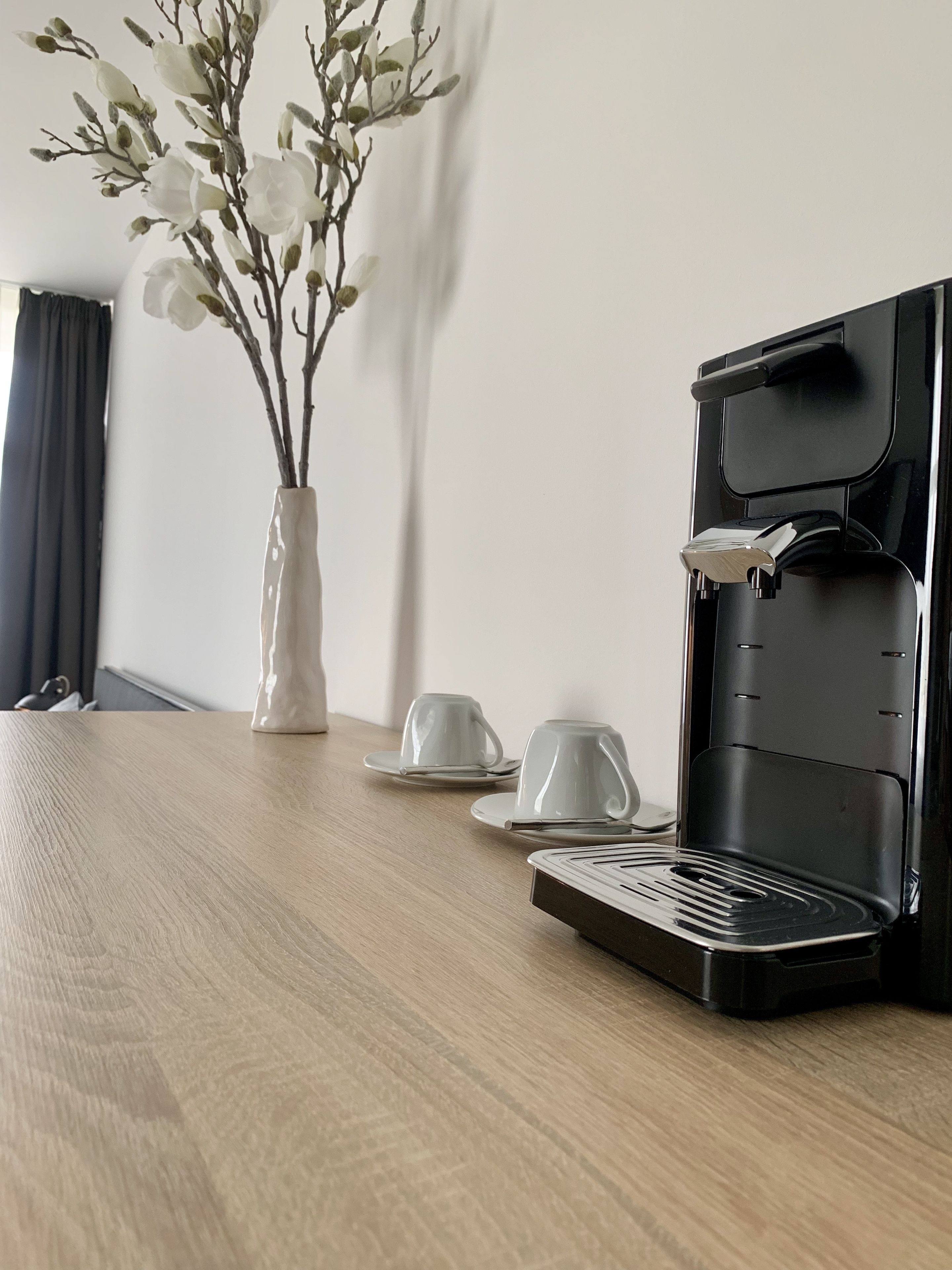 Uma máquina de café sobre uma mesa de madeira, com duas chávenas e pires brancos, um vaso com flores brancas e uma janela com cortinas.