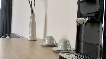 Uma máquina de café sobre uma mesa de madeira, com duas chávenas e pires brancos, um vaso com flores brancas e uma janela com cortinas.