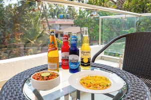 Uma mesa de vidro com garrafas de cerveja e uma tigela de comida em uma varanda, com vista para árvores e edifícios.