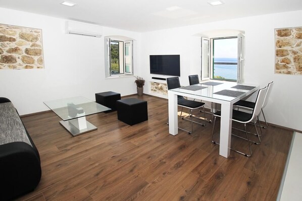 Quarto moderno e bem iluminado com mesa de jantar de vidro, cadeiras pretas, sofá e televisão na parede.