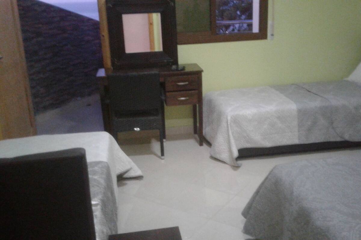 Quarto pequeno e simples com duas camas de solteiro, uma escrivaninha com cadeira e uma janela.