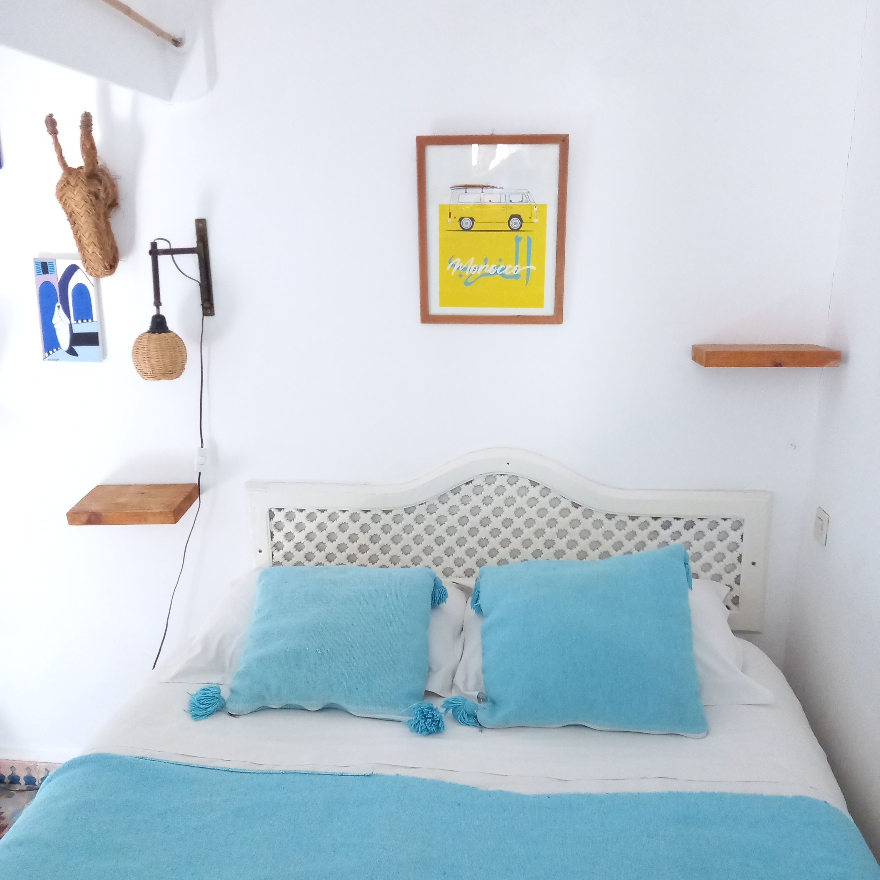 Uma cama com cabeceira branca, duas almofadas azuis e um cobertor azul. Há um quadro na parede com uma carrinha amarela.