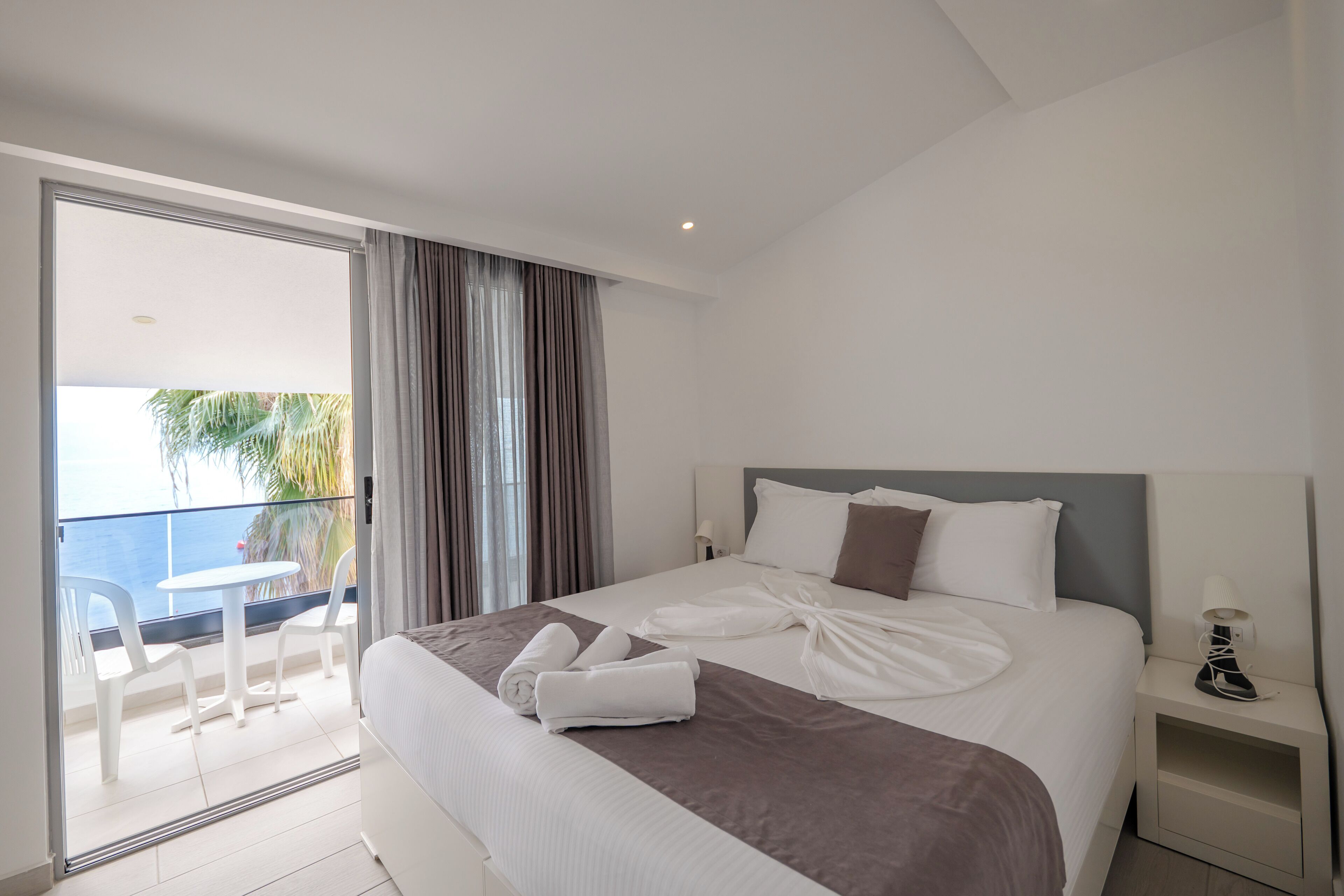 Quarto de hotel com uma cama grande, uma mesa de cabeceira, uma luminária e vista para uma varanda com cadeiras e uma mesa.