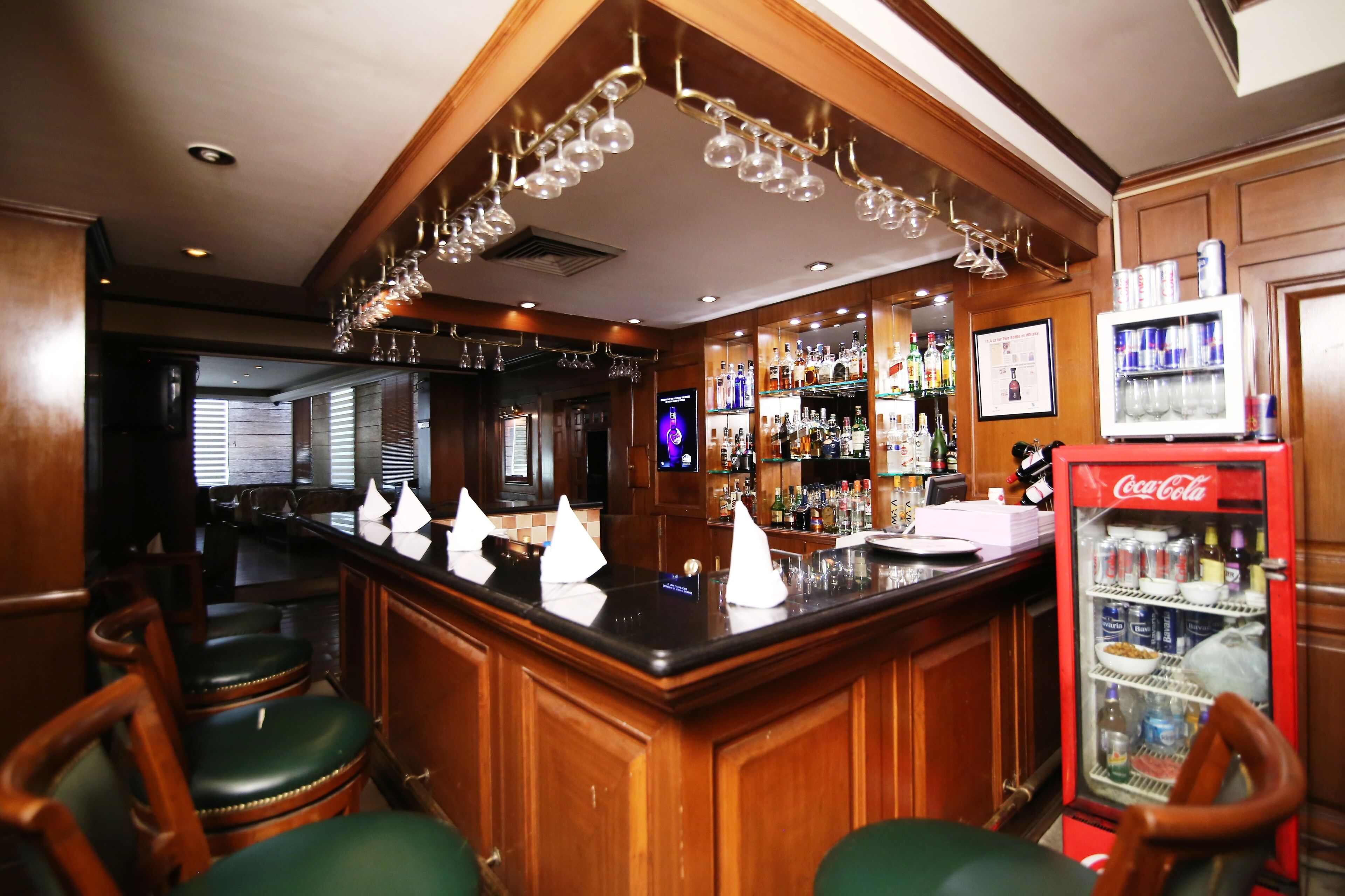 Uma área de bar com balcão de madeira, banquetas verdes, luminárias pendentes e um refrigerador de Coca-Cola.