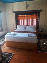 Cama bem arrumada com lençóis e almofadas brancos, cabeceira de madeira, mesa de cabeceira com abajur e tapete estampado no chão de madeira.