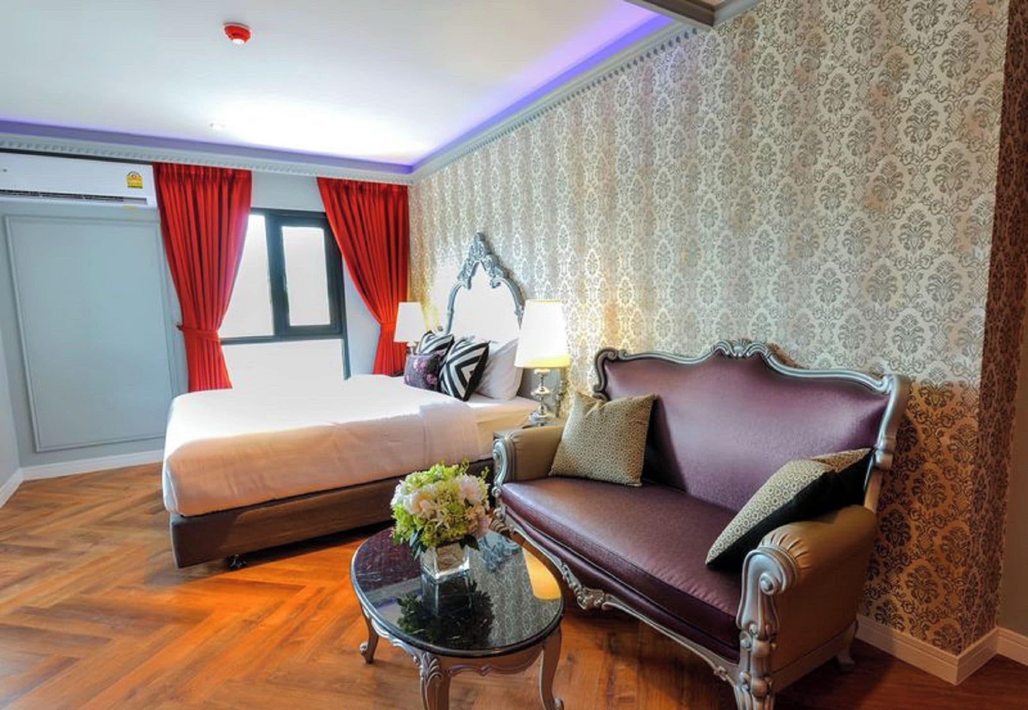 Quarto de hotel com cama, sofá, mesa com flores e papel de parede estampado.