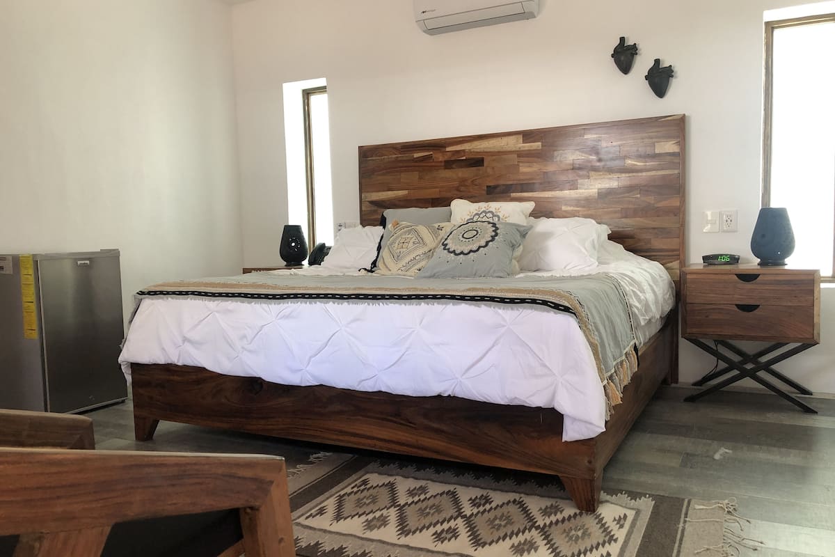 Quarto com cabeceira de madeira, cama com roupa de cama branca, mesa de cabeceira com relógio e ar-condicionado fixado na parede.