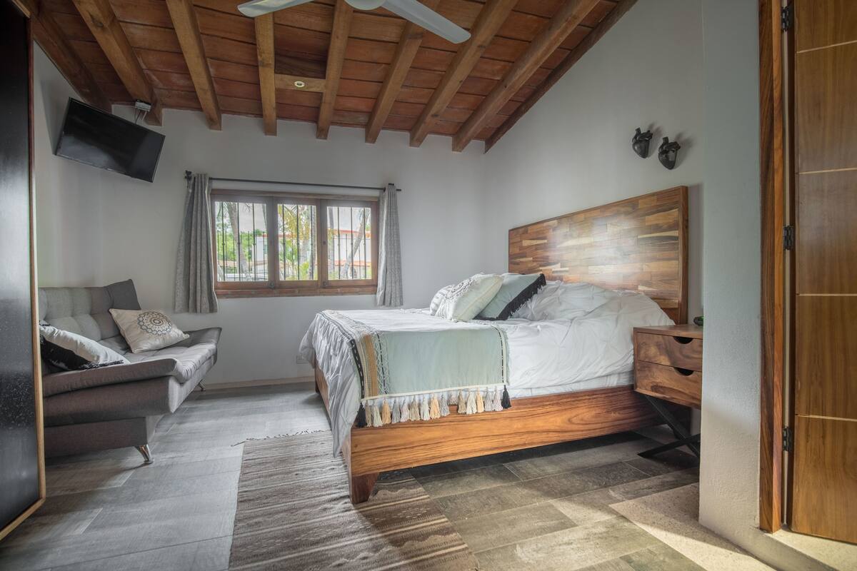 Quarto com cama de madeira, um sofá, televisão fixada na parede e janela com cortinas.