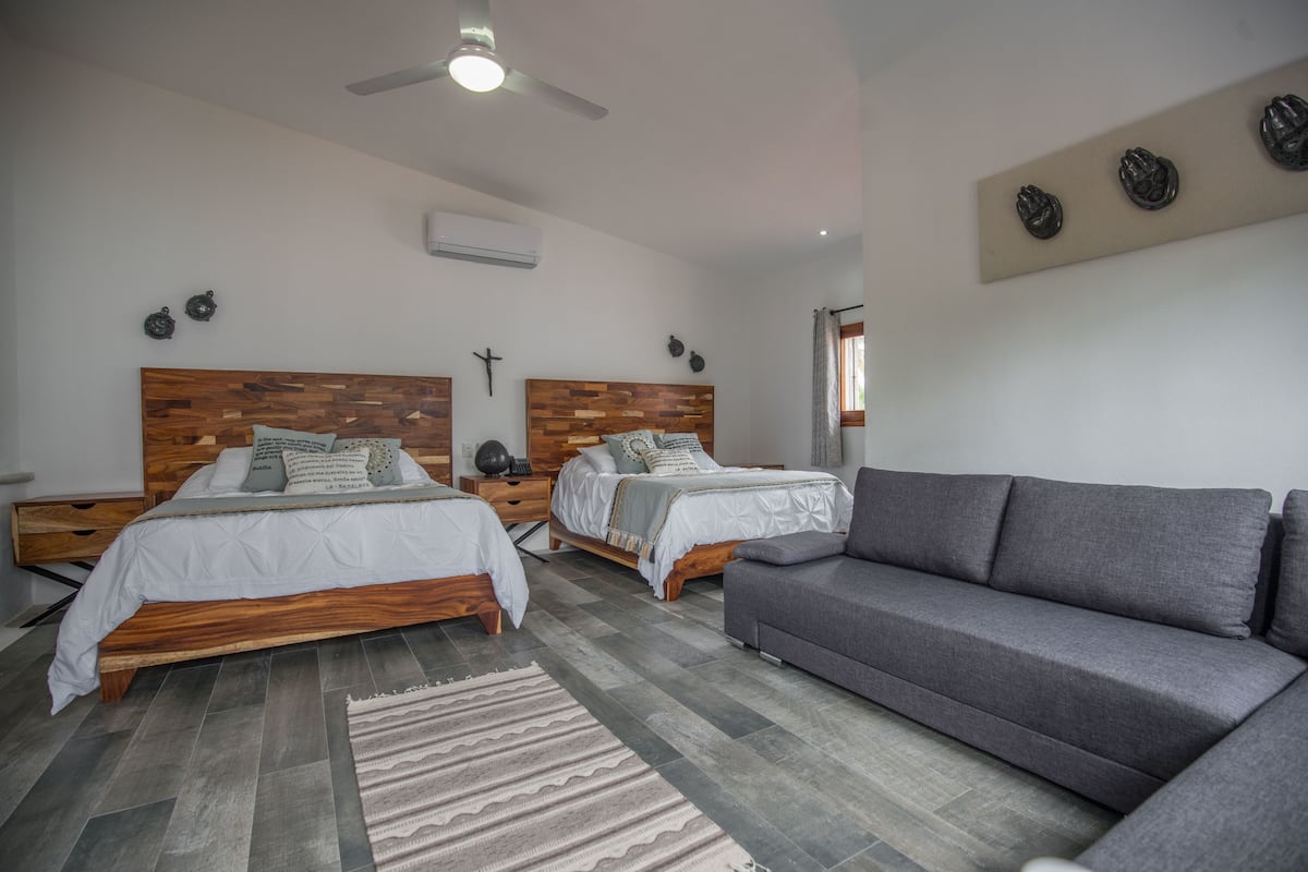 Quarto com duas camas, um sofá cinzento e um ventilador de teto.