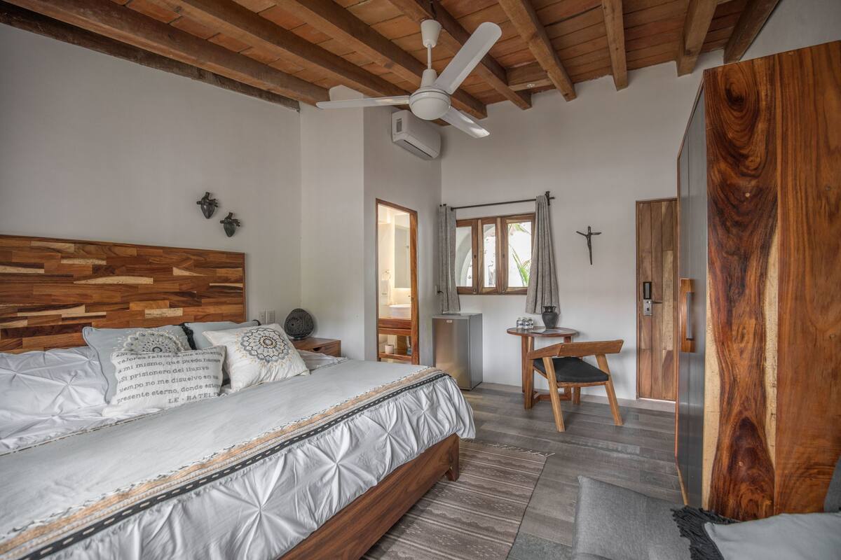 Quarto com cabeceira de madeira, cama com roupa de cama estampada, escrivaninha com cadeira e uma cruz na parede.