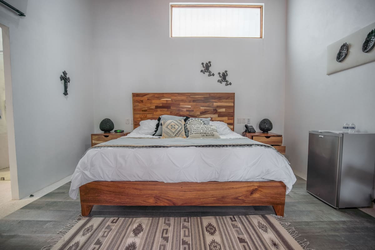 Quarto com cabeceira de madeira, colcha branca, almofadas decorativas, mesa de cabeceira pequena com abajur e um frigorífico.