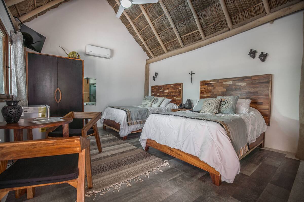 Quarto com duas camas, cabeceiras de madeira, uma escrivaninha com cadeira, uma mesa pequena e teto de palha.