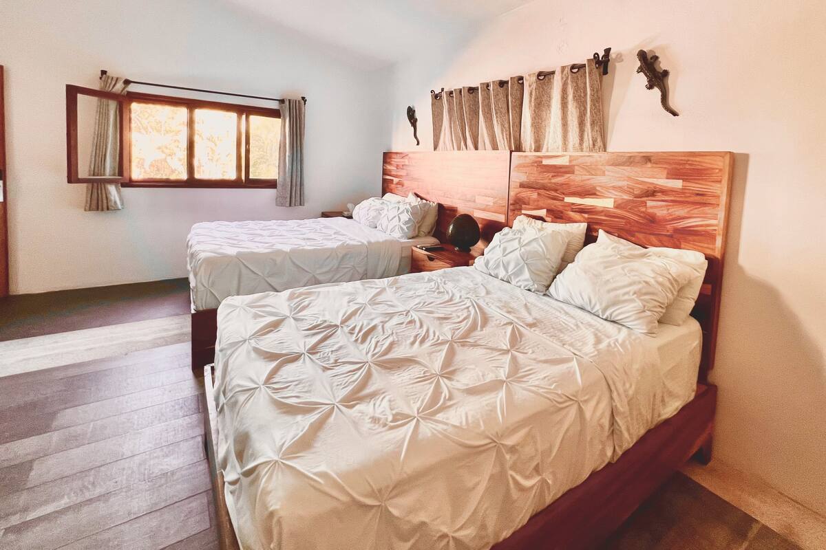 Quarto de hotel com duas camas, cabeceiras de madeira, uma janela grande com cortinas e piso de madeira.