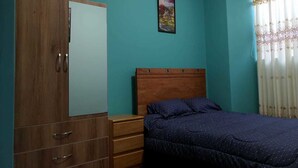 Quarto com paredes verde-água, um guarda-roupa de madeira, uma cama com colcha azul e uma mesa de cabeceira.