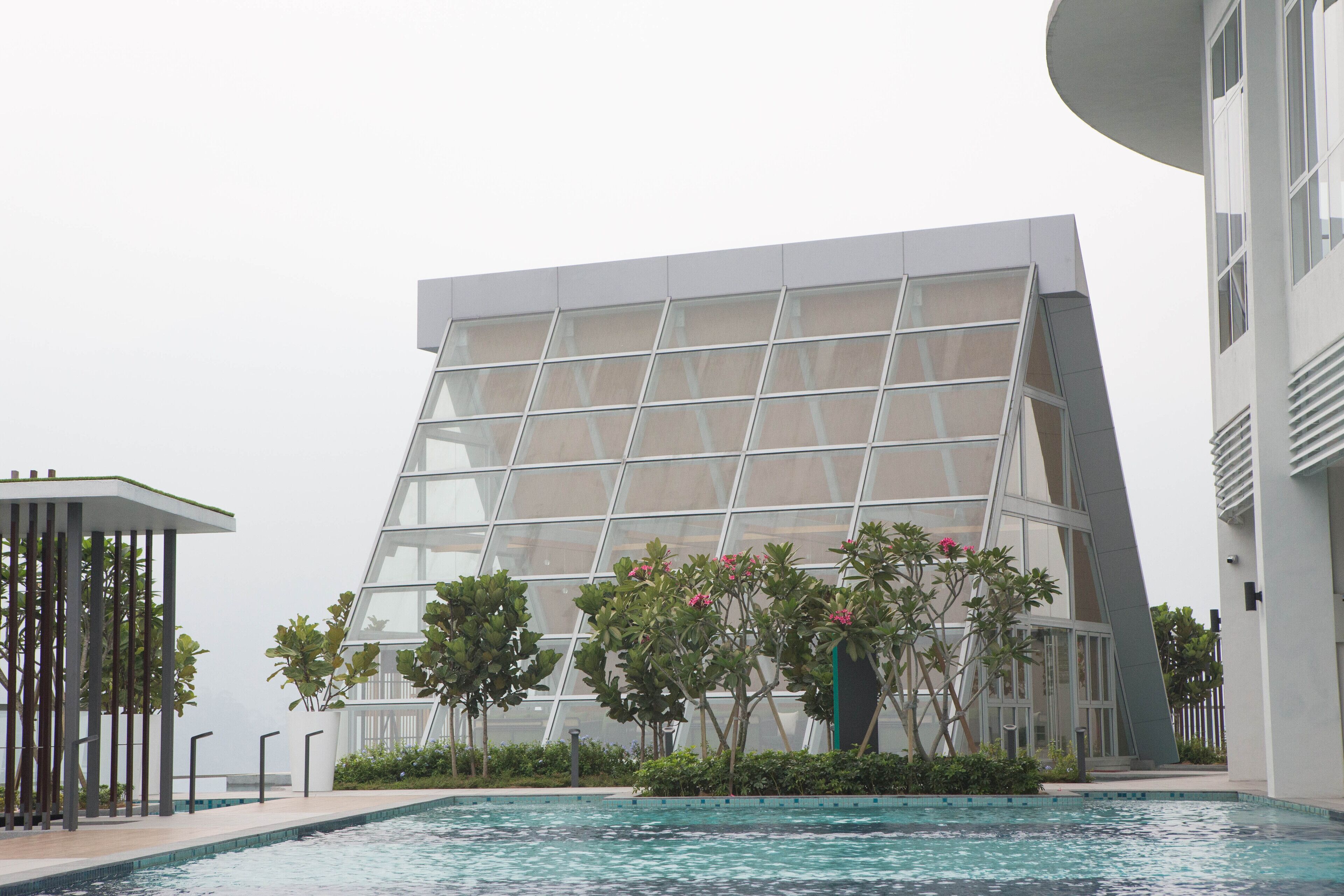 Um edifício moderno com fachada de vidro, piscina e vegetação ao redor.