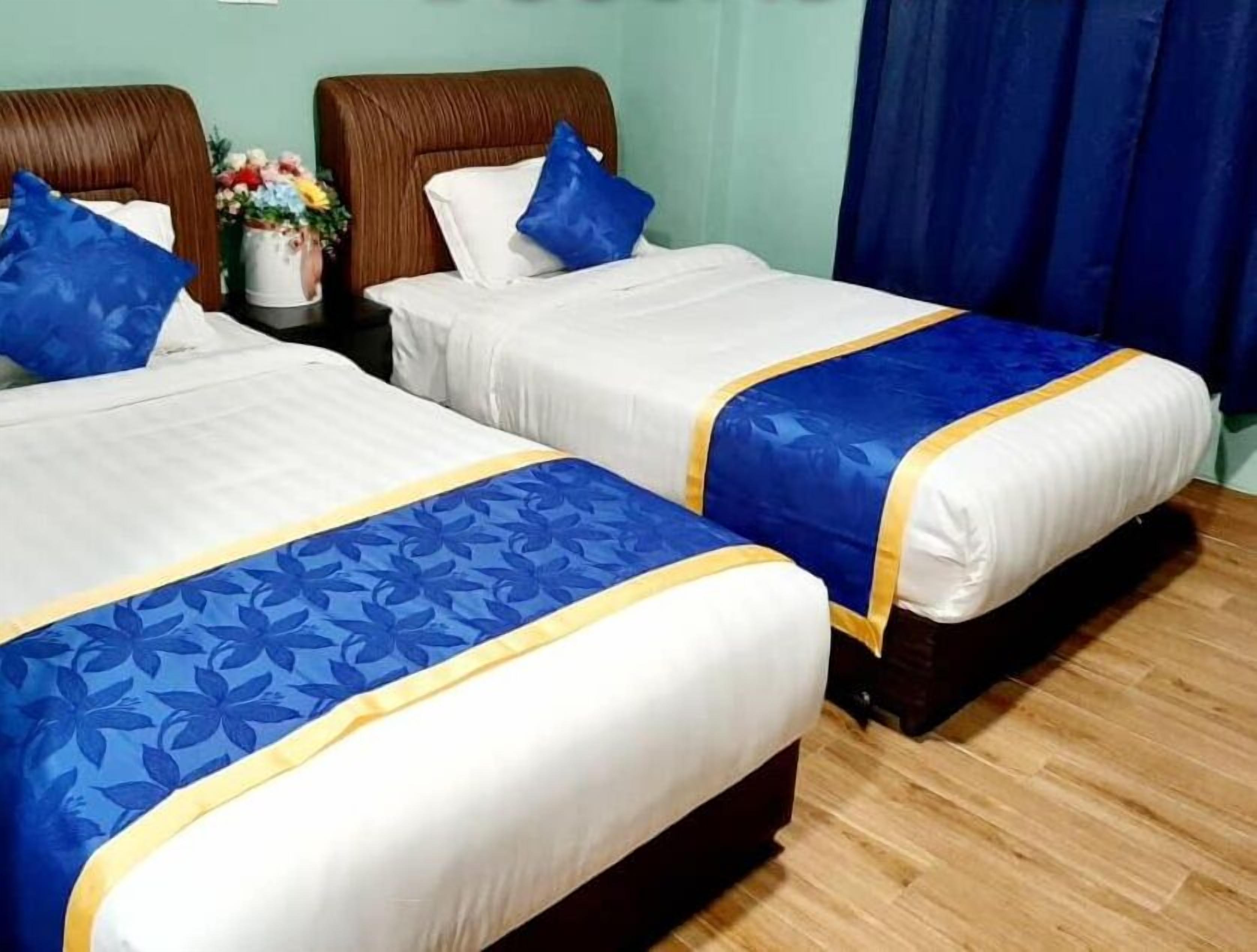 Duas camas com roupa de cama azul e branca, um cabeceira de madeira, uma mesa de cabeceira com um vaso de flores e uma cortina azul.